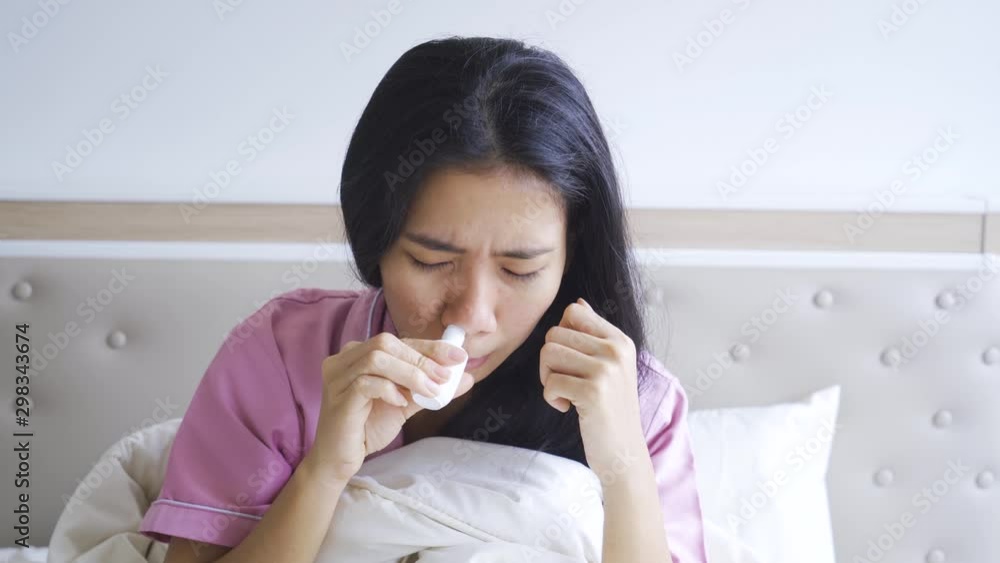Sick woman sniffing nasal spray while sitting on the bed in bedroom at ...