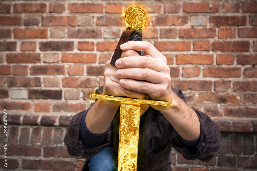 Warrior kneels in a hooded cloak and holds a shining golden sword in ...