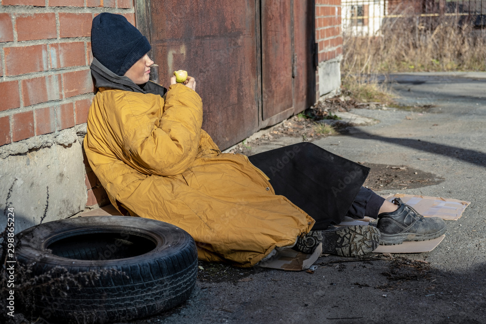 Homeless woman in a yellow old ragged jacket and blue hat is sitting on ...