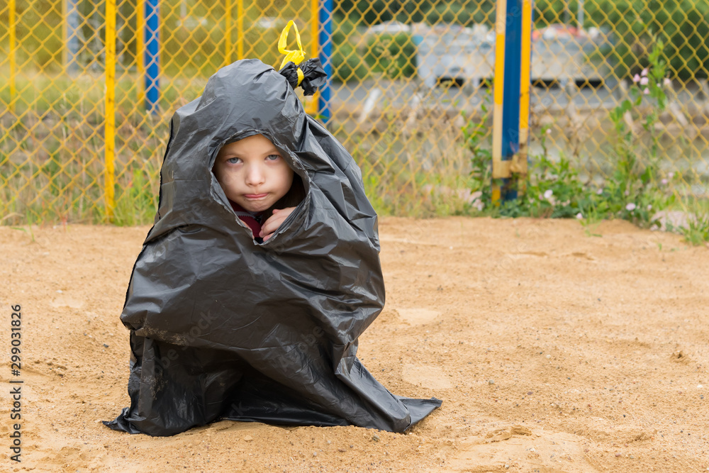 sad child sitting in a black garbage bag, on the background of a yellow ...