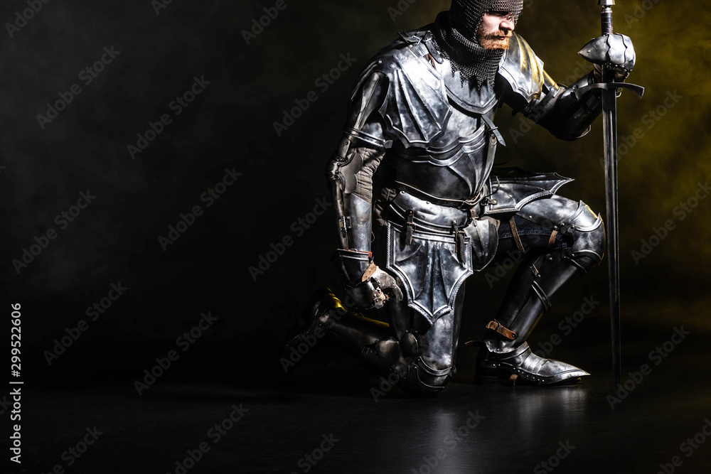 handsome knight in armor holding sword and bend knee on black ...