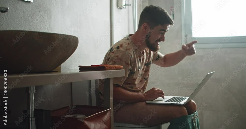 Bearded young man working on laptop computer sitting on toilet in the ...