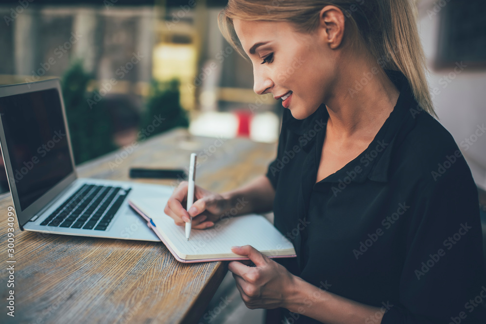 Side view of positive skilled female student making notes of ...