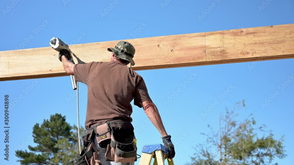 Zooming in on a male contractor on a yellow ladder, using a nail gun on ...