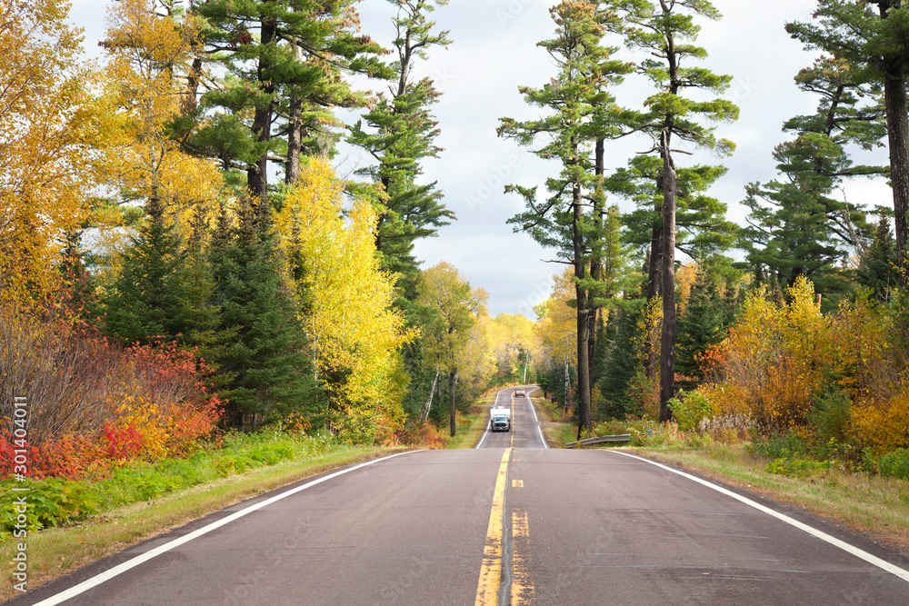 Camper and pickup truck drive below tall pines and autumn color on ...