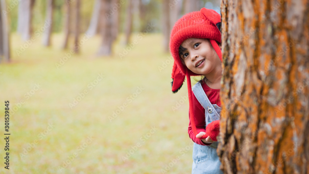 Adorable smiling asian little girl wear a red hat hide herself behind ...