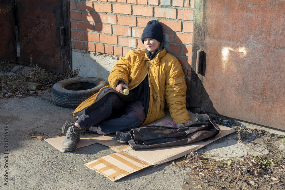 Homeless woman in a yellow old ragged jacket and blue hat is sitting on ...
