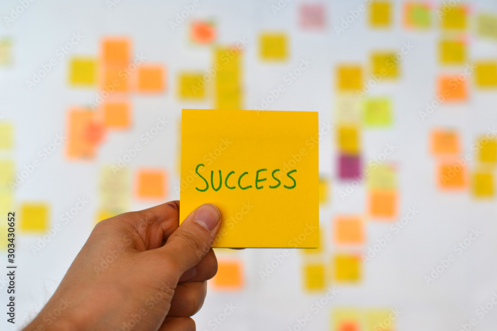 A hand is holding a orange success sticker and there is a Kanban board ...