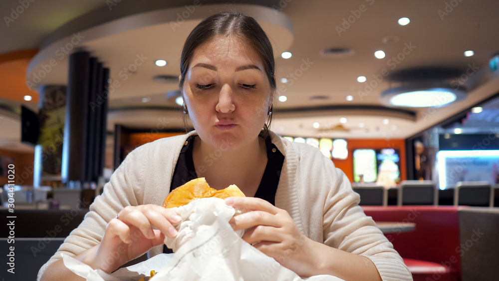 Junk food concept. Young brunette woman is eating burger in fast food ...