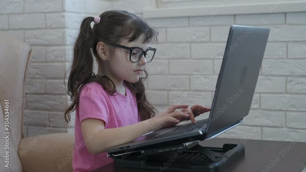 The child is at the computer. Cute little girl in glasses is typing on ...