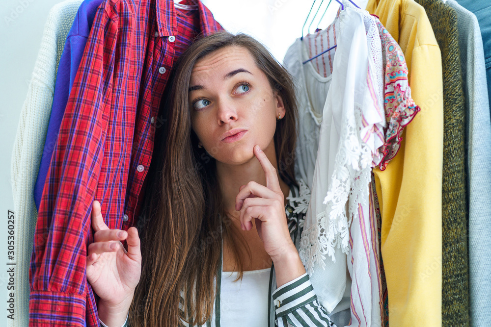 Thinking female standing near wardrobe rack full of clothes on hangers ...