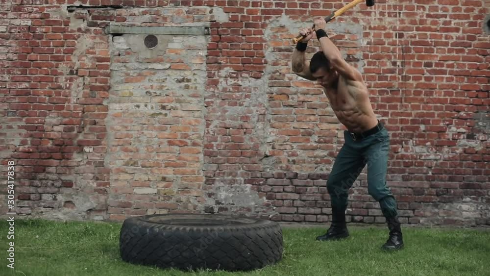 Strong muscular shirtless young man hitting a tire with a sledgehammer ...