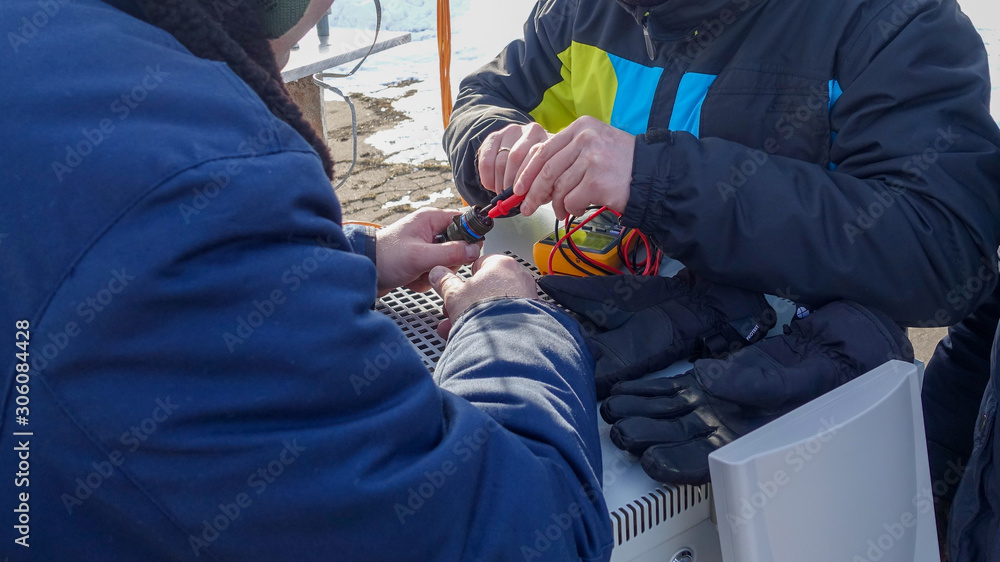 Two men in warm winter clothes use a digital multimeter to check the ...