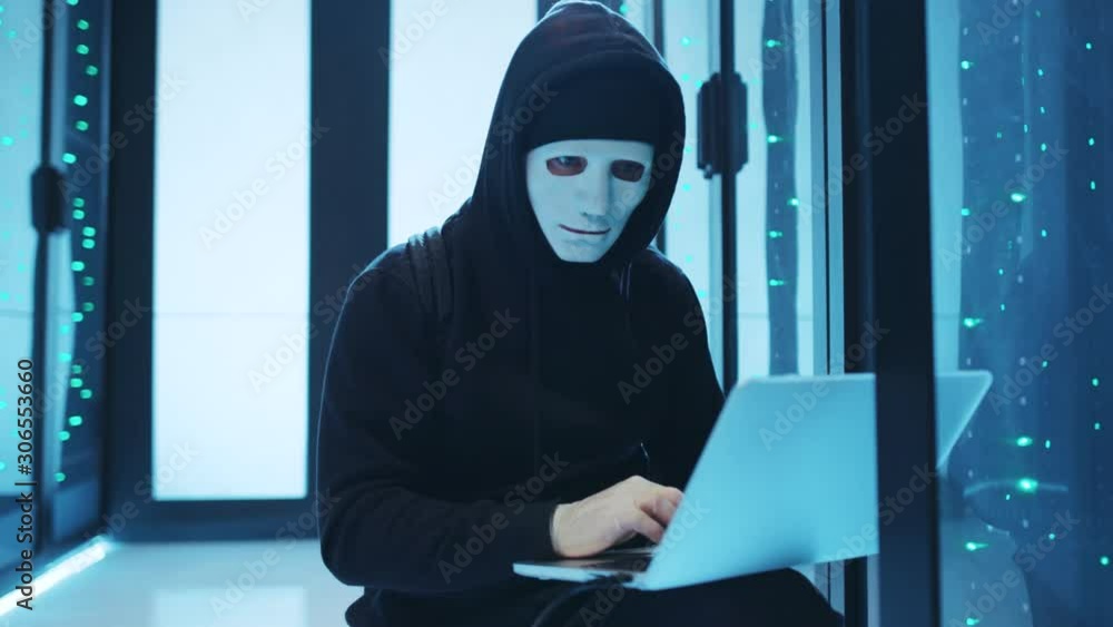 Masked hacker in black hoodie typing code on laptop using malicious software to steal important ...