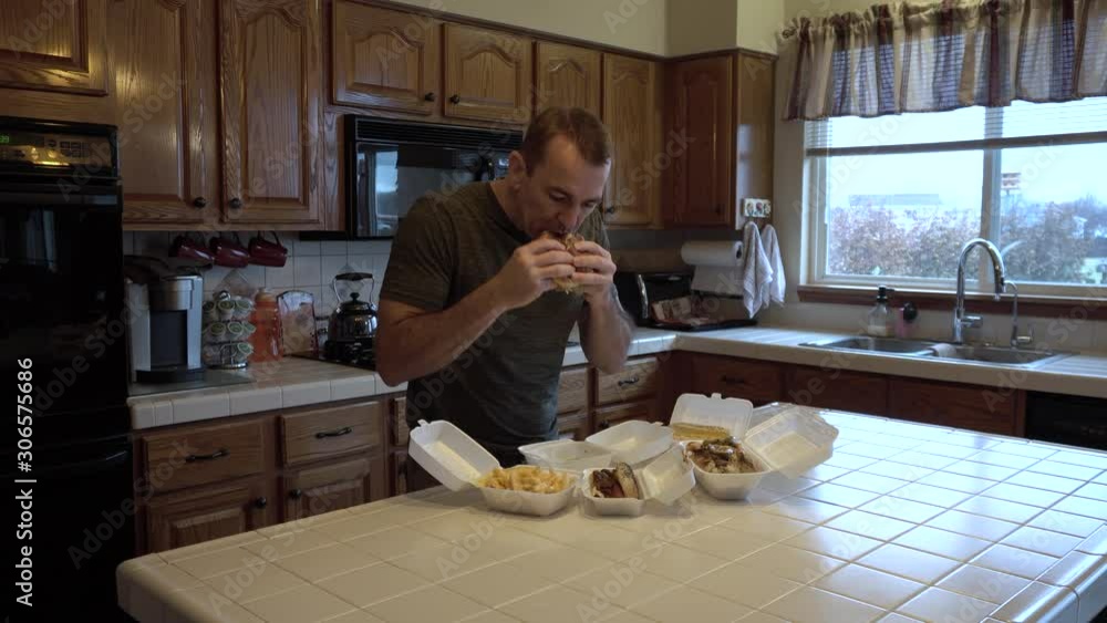 Man standing eating a hamburger in the kitchen, overeating, unhealthy ...