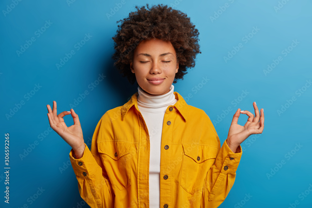 Relieved calm dark skinned woman closes eyes, makes mudra gesture ...