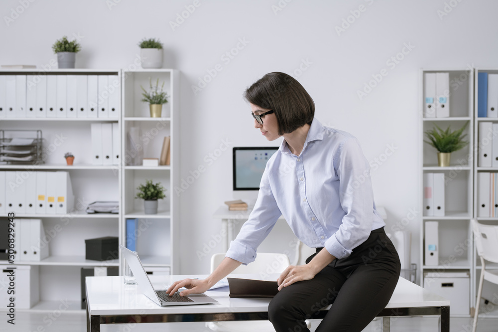 Young elegant businesswoman sitting on desk and slighlty bending over ...