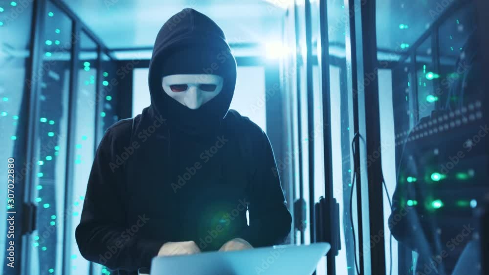Concentrated masked hacker in hoodie coding malicious program on laptop ...