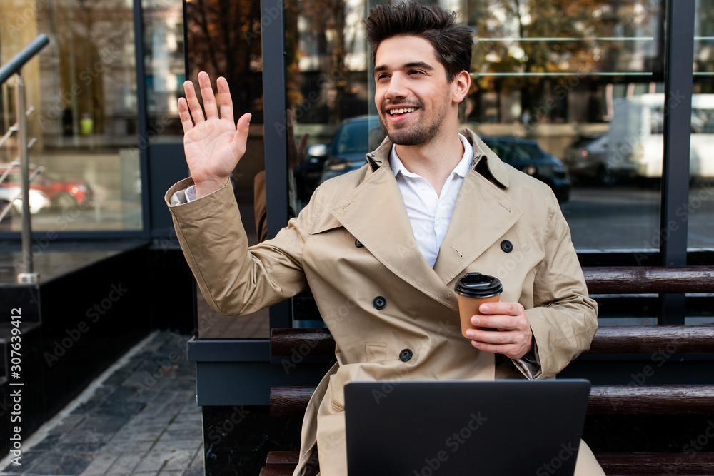 Young attractive smiling man in trench coat with laptop and coffee ...