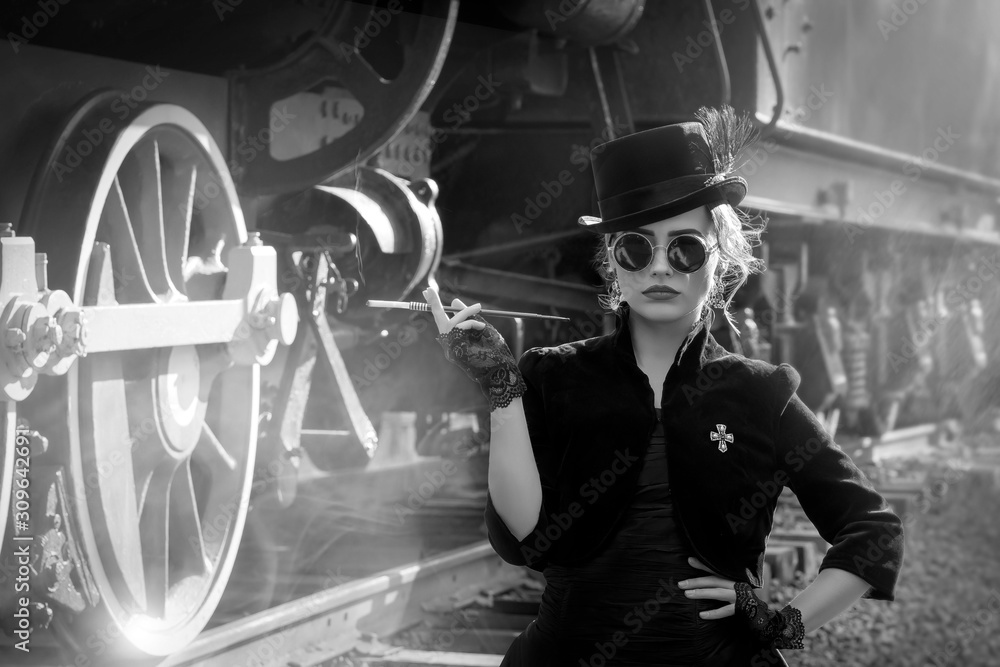 Steampunk girl in a black dress and hat near an old steam locomotive ...