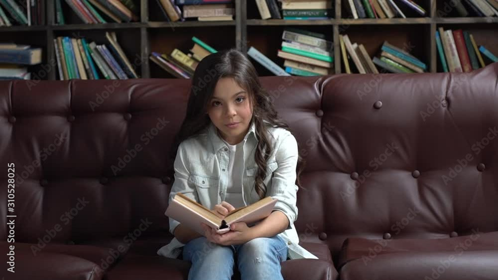 Little girl sitting in library reading book looking camera curious ...