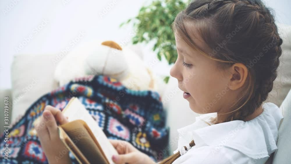 Vidéo Stock Cute little girl sitting with her legs curled up, reading ...