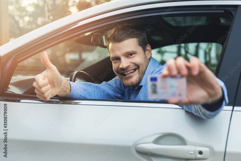 young smiling man sitting in the car and showing his new driver license ...
