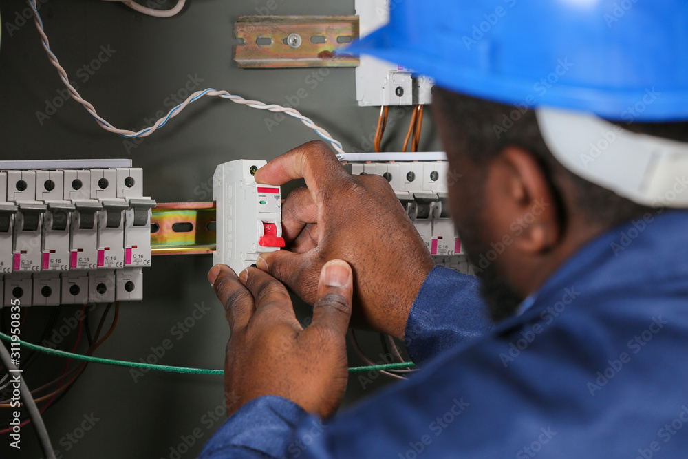 African-American electrician performing wiring in distribution board ...