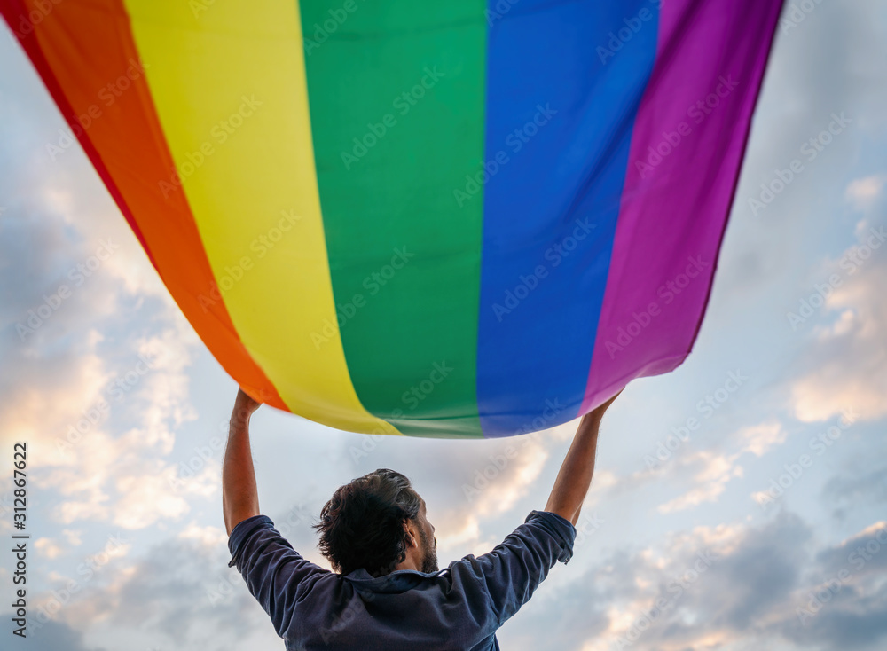 Cheerful guy with a rainbow flag on the beach. Young man holding a ...