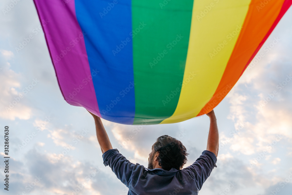 Cheerful guy with a rainbow flag on the beach. Young man holding a ...