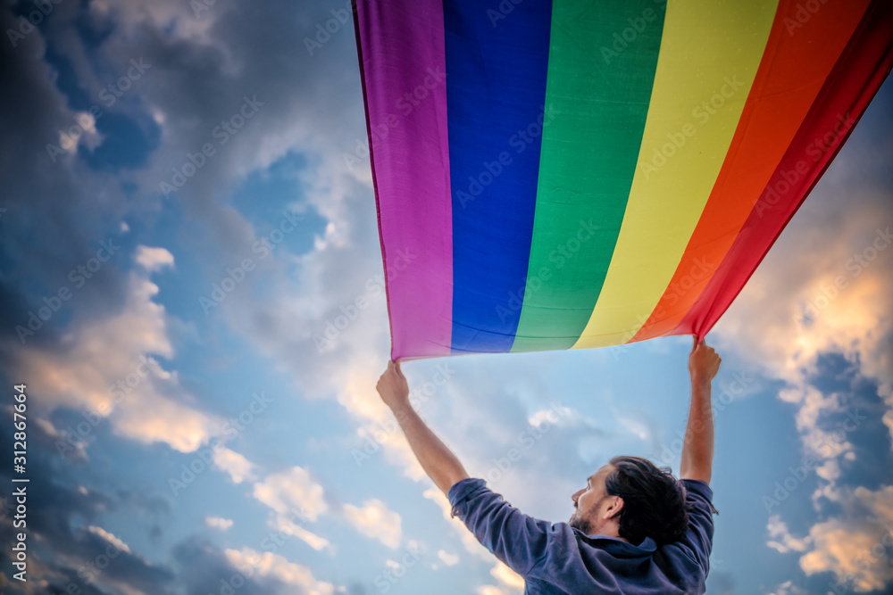 Cheerful guy with a rainbow flag on the beach. Young man holding a ...