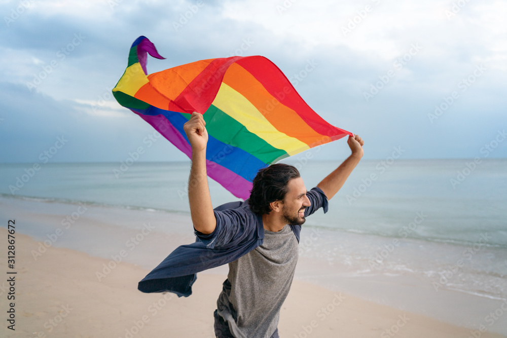 Cheerful guy with a rainbow flag on the beach. Young man holding a ...
