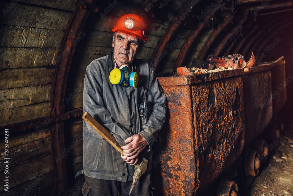 An elderly man dressed in work overalls and a hard hat is standing near ...
