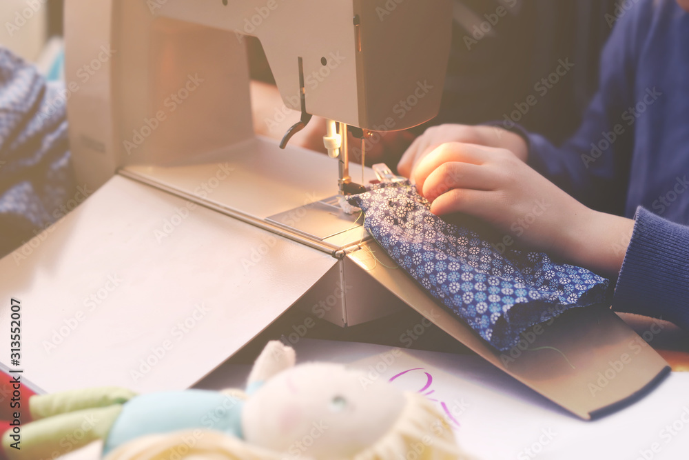 Enthusiastic young girl sews a toy dress for her doll at a compact ...