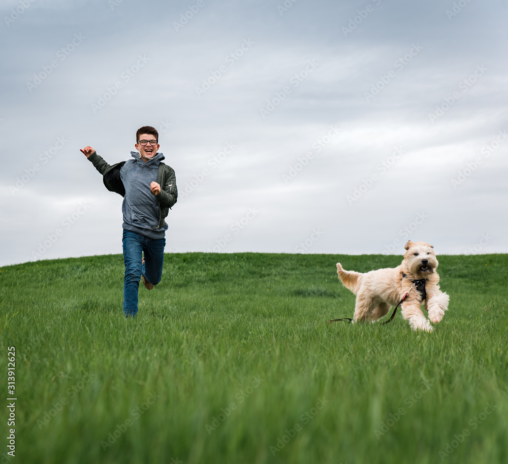 Teenage boy running across a grassy field with his dog on cloudy day ...