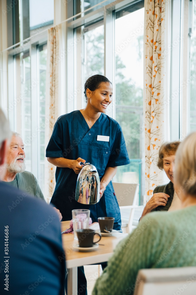 Smiling female nurse serving drink to senior women and men at dining ...
