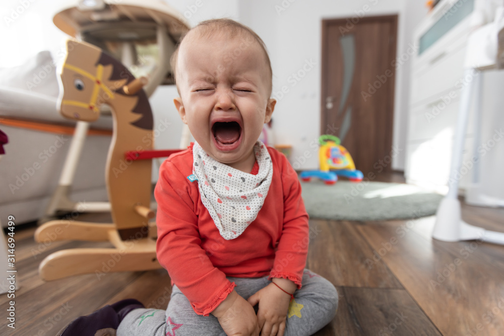 Little girl sitting on the floor at home and crying. One year old baby ...
