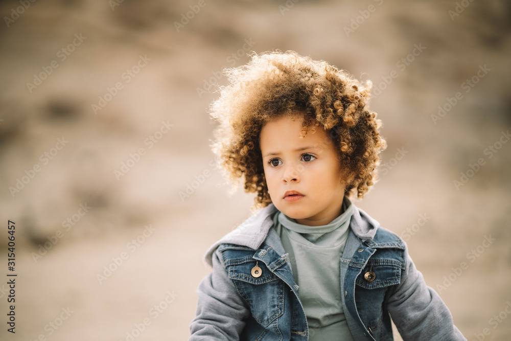 Adorable curly ethnic child dressed in casual clothes on beige blurred ...