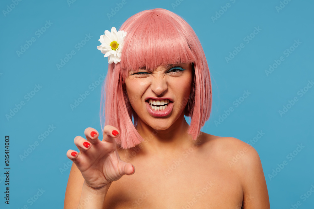 Playful young pretty lady wearing chamomile in her short pink hair while posing over blue ...
