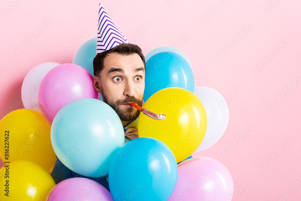 handsome man in party cap with party blower in mouth near balloons on ...
