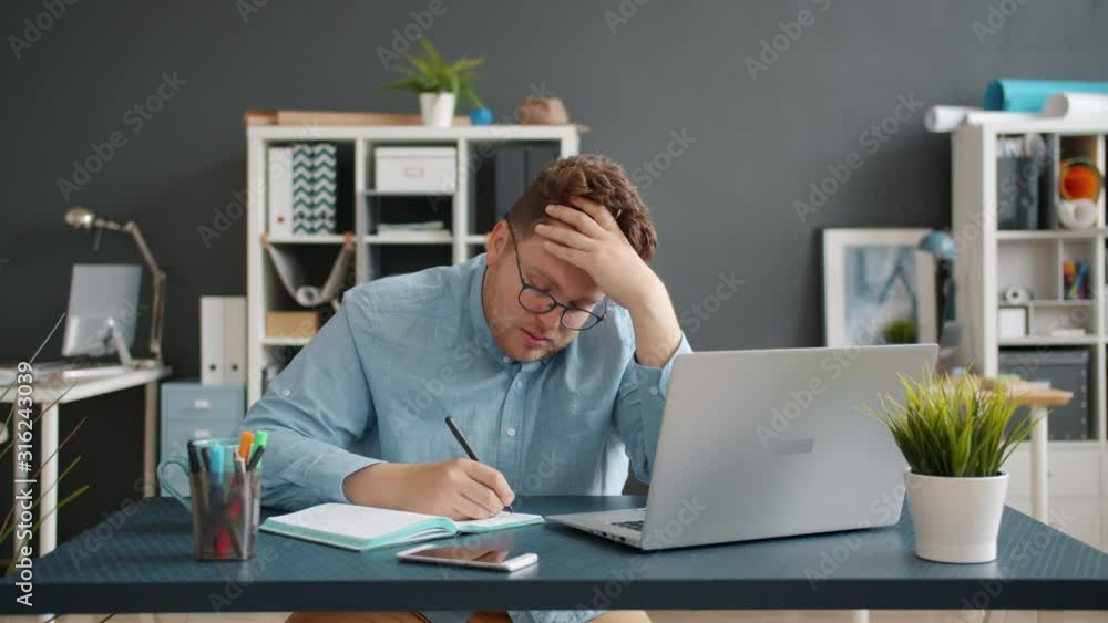 Exhausted young man is writing and working with computer indoors at ...