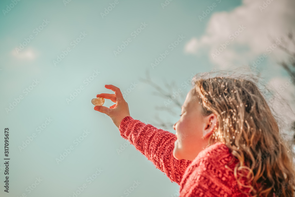 Beautiful blonde girl holding Heart shaped stone in hands, blue sky in ...