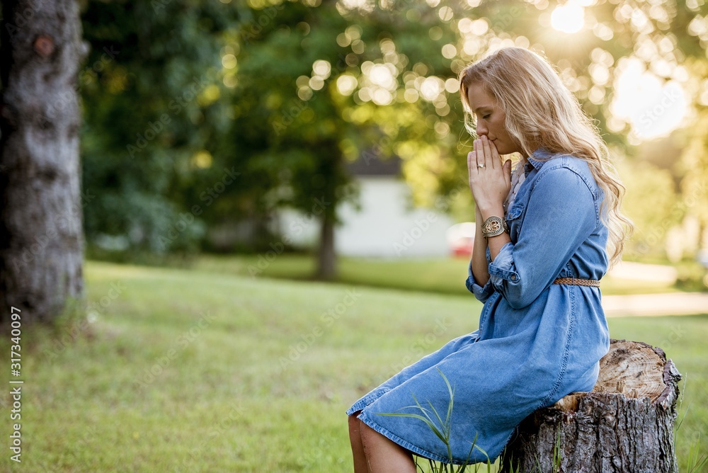 Blonde woman sitting on a tree stump and praying in a garden under ...