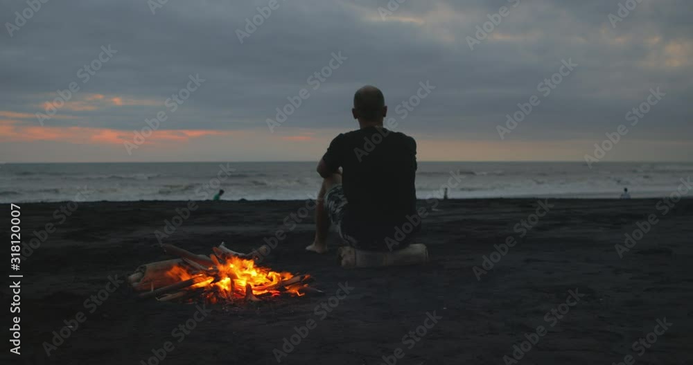 Young guy sits at the campfire on the beach. Young urban man near fire ...