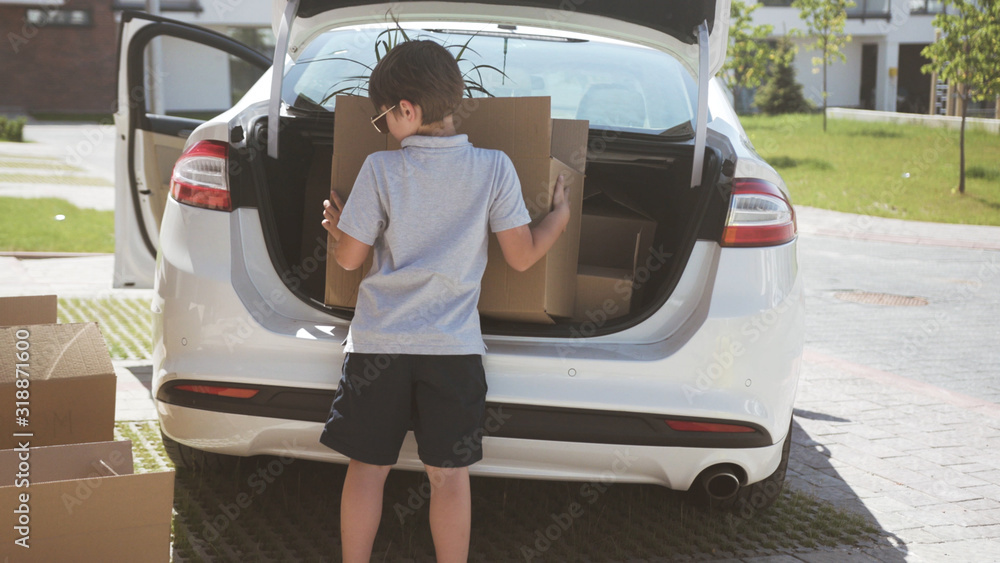 Little strong boy loading heavy boxes with goods into a car trunk ...