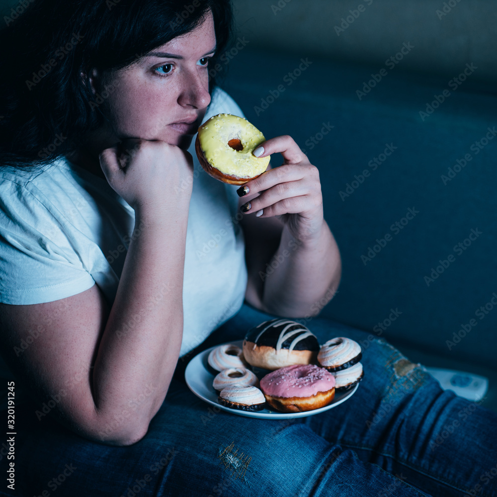 Overweight depressed woman laying on sofa eating sugary food watching ...