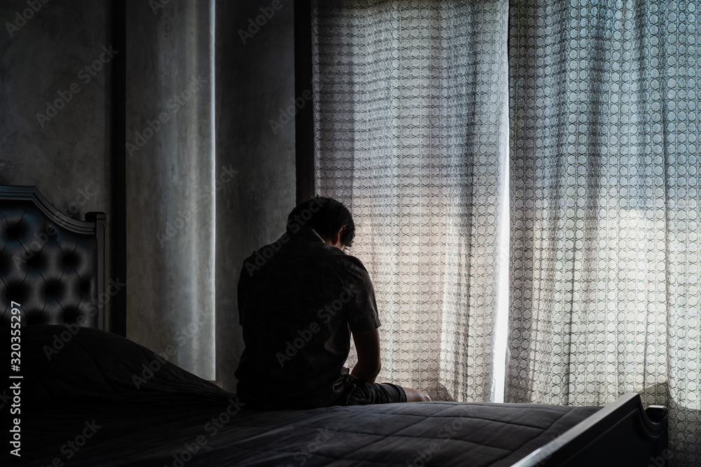 Lonely young man feeling depressed and stressed sitting head in hands in the dark bedroom ...