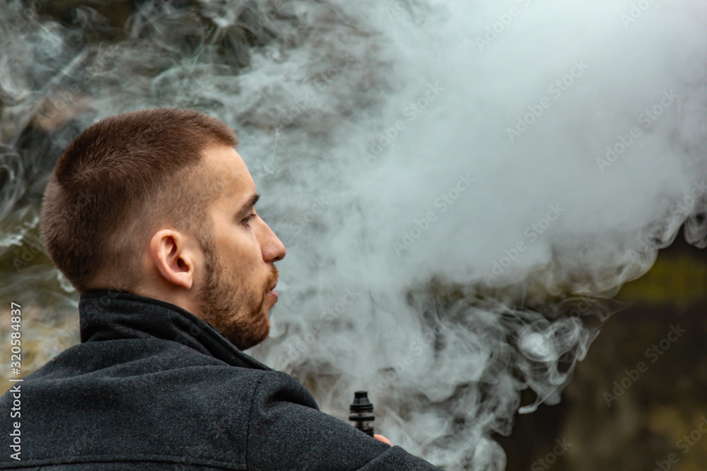 A guy with a beard sits on the lake and smokes an electronic cigarette ...