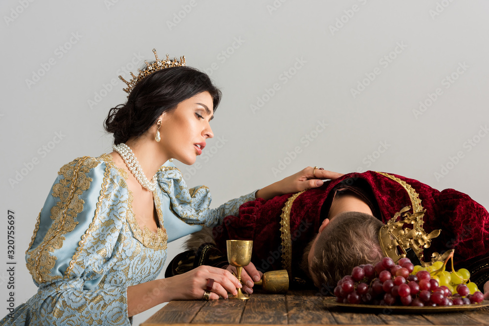 sad queen with crown looking at killed king isolated on grey Stock ...