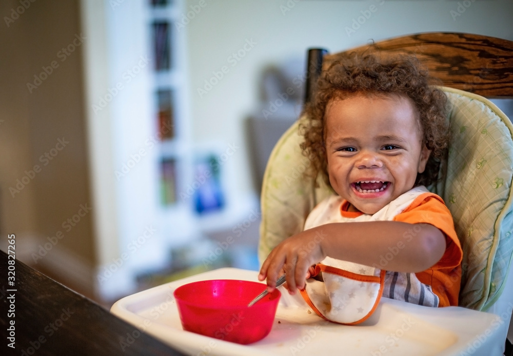Cute Laughing diverse Child sitting in his high chair eating a meal ...